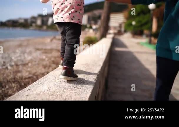 Small baby runs along a stone curb holding his mother hand. Back view ...