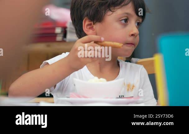 Boy eating breadstick at table, holding snack, relaxed expression ...