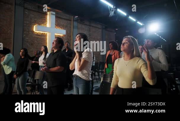 People worshiping together in church, raising hands in prayer, illuminated cross in background ...