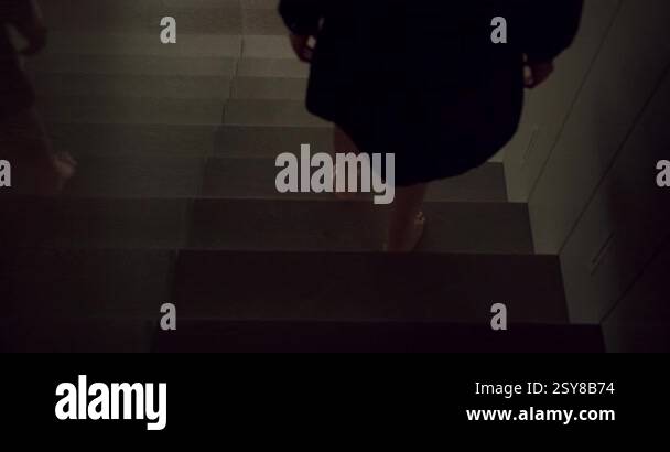 A shadowy, unidentifiable person walks down dark, neatly tiled stairs. The image evokes a sense ...