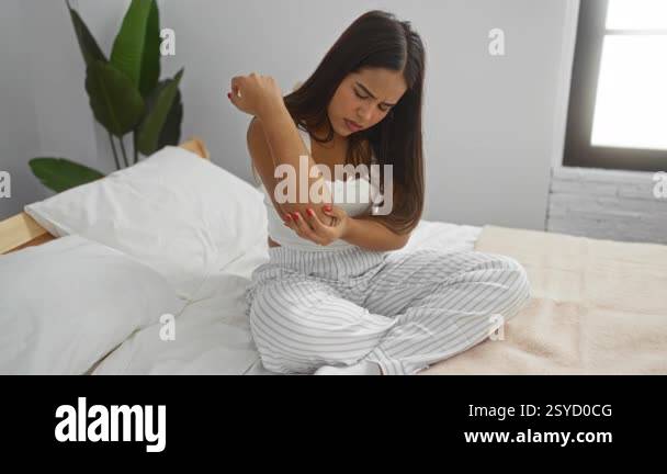 Woman sitting on bed in bedroom examining elbow with a concerned look ...
