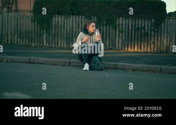 Young woman sitting on curb in residential area drinking water. Female ...
