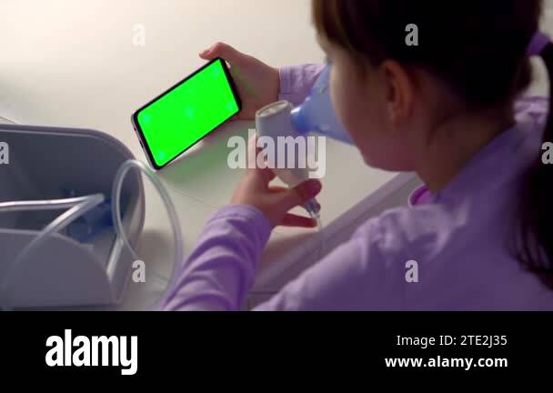 Little girl is sick, makes inhalation with nebulizer and looks at phone ...