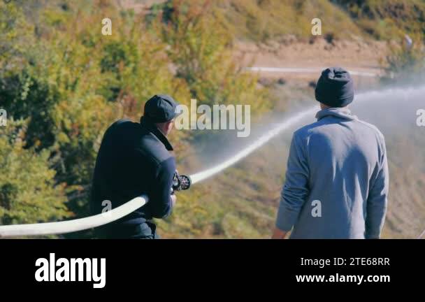 A two men holds a fire hose and waters the object under high pressure ...