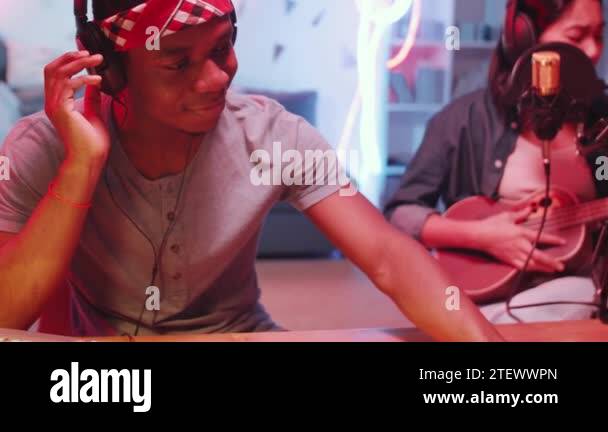 Young Afro-American man in headphones sitting at desk and using mixing console while recording ...