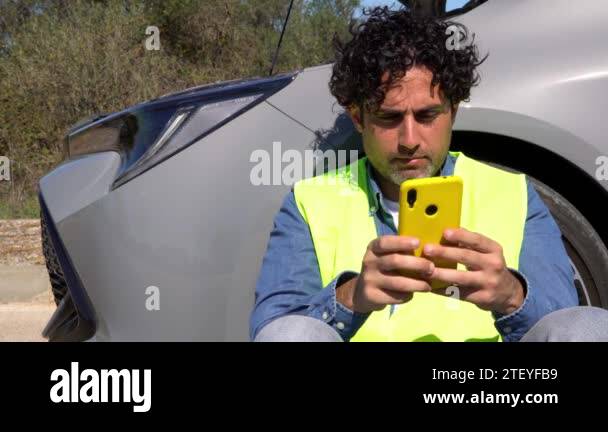 Angry man texting for assistance, sitting on asphalt near broken car with popped up hood at the ...