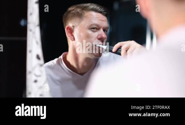 Focused handsome young man electric brushing teeth in front of mirror ...