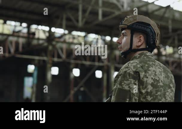 Serious military man in helmet and uniform standing in ruined plant ...
