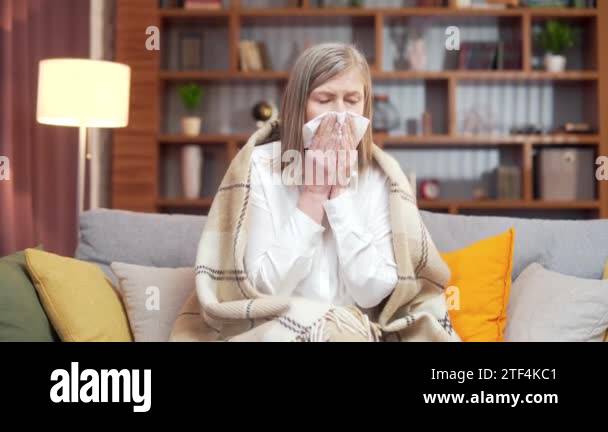 Gray hair aged elderly woman with flu symptoms sitting on couch blowing ...
