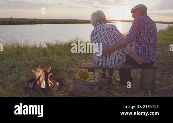 Rear view at two old hugging spouses sitting at lake beach enjoying ...