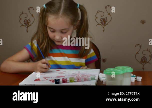 A girl of 7-8 years old sits at a table and draws a picture with paints ...