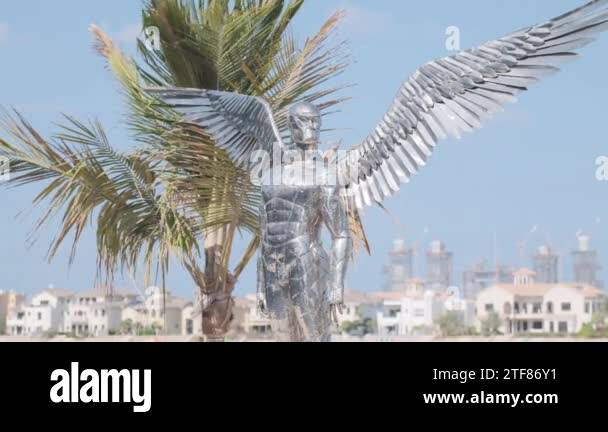 Winged man metal statue is standing in front of the palm on the street ...