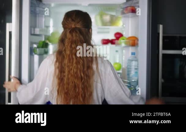 Young woman taking vegetables from fridge and has fun singing and ...