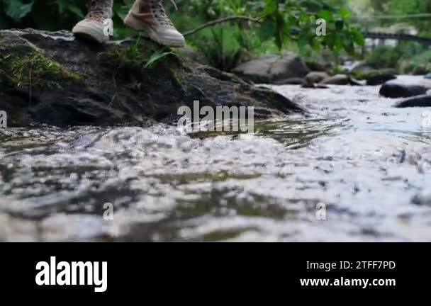 Female traveler walks on a rock across a stream in the forest. View of ...