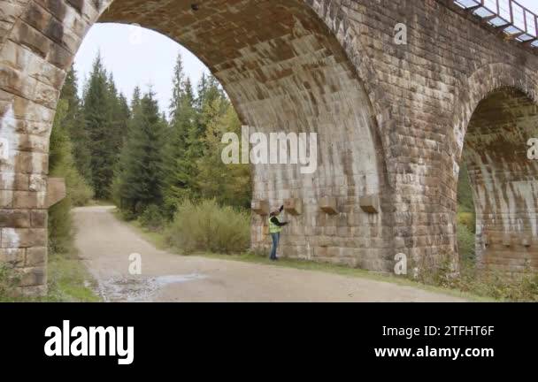 A senior gray-haired engineer stands under a very old bridge and takes ...