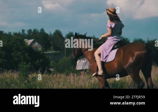 Young woman in a summer dress and straw hat riding a horse in a rural ...