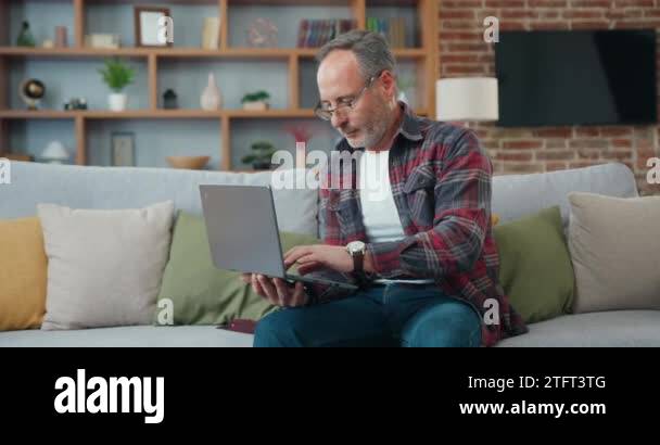 Handsome mature man working on laptop while sitting on a sofa in living ...
