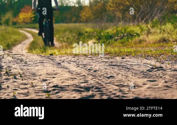 Young guy rides a bicycle along a dusty path in the forest at sunset. A ...
