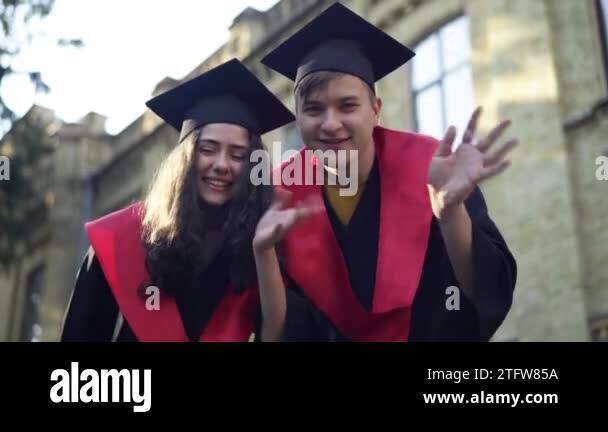 Joyful couple in graduation togas and mortarboard caps waving smiling ...