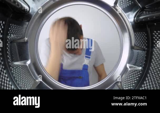 a home appliance repairman in blue uniform sticks his head into drum of ...