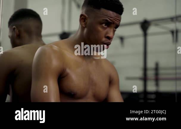 Shirtless muscular young black man resting and breathing with reflection in mirror at the gym ...
