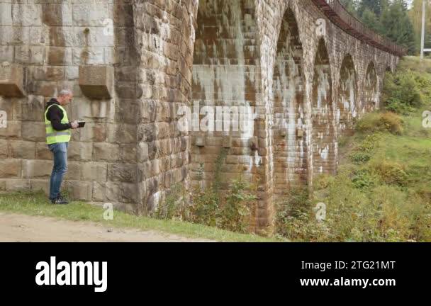 A senior gray-haired engineer stands under a very old railway bridge ...