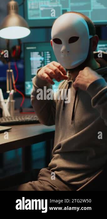 Vertical video: Masked scammer hacking computer network late at night, working on laptop and ...