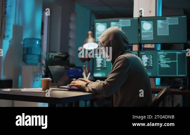 Masked data thief hacking computer network at night, working on laptop and multiple monitors to ...