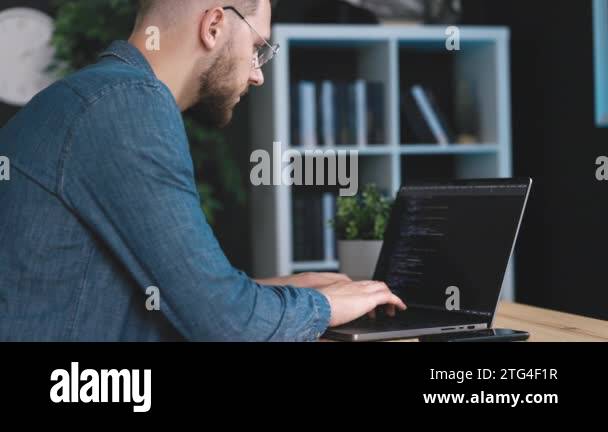 Man in denim shirt coding software at laptop, desk with computer ...