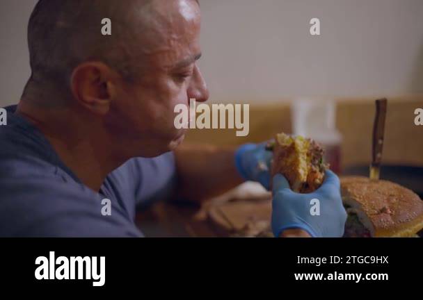 Adult bald man eating a piece of juicy burger with gloves, enjoying ...