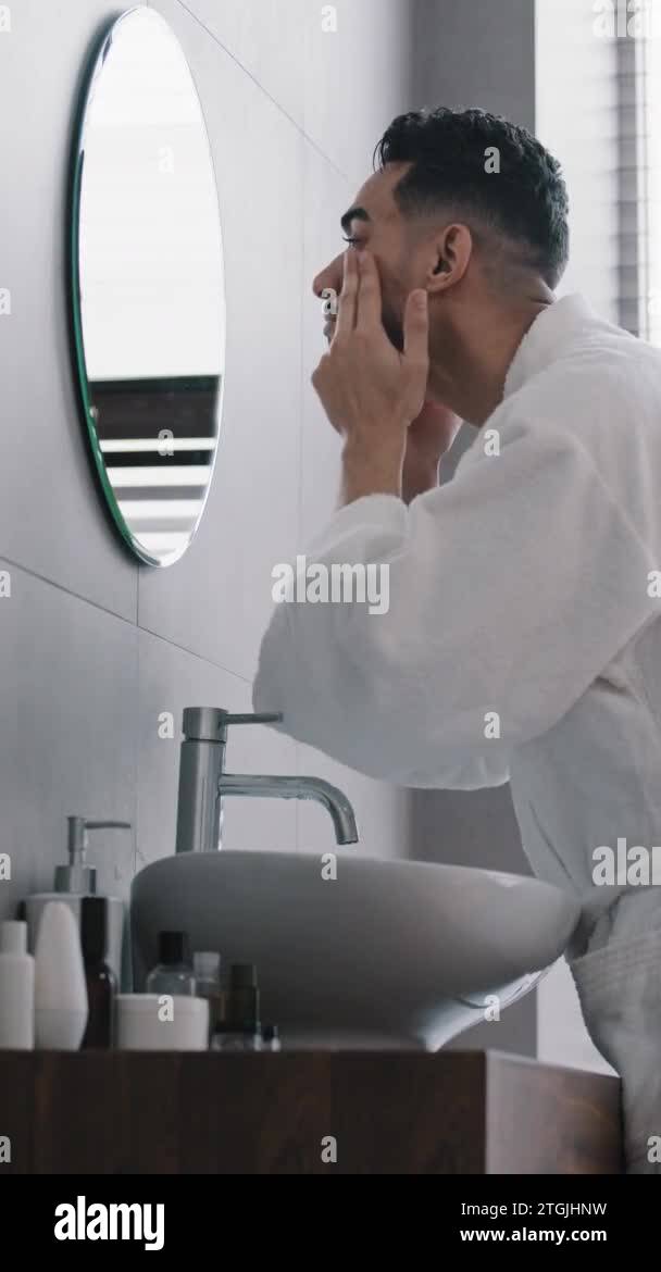 Vertical view Arab Spaniard Indian man bearded guy in bathrobe looks at reflection in mirror in ...