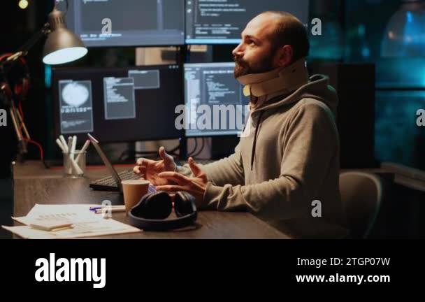 Adult programmer with neck collar foam coding html script, using laptop and multiple monitors at ...