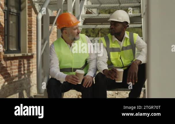 Smiling multicultural builders in hardhats looking at camera with smile while sitting together ...