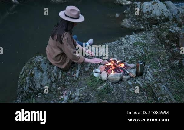 Young woman in hat sitting near campfire in forest. Hiking girl in hat looks at bonfire and ...