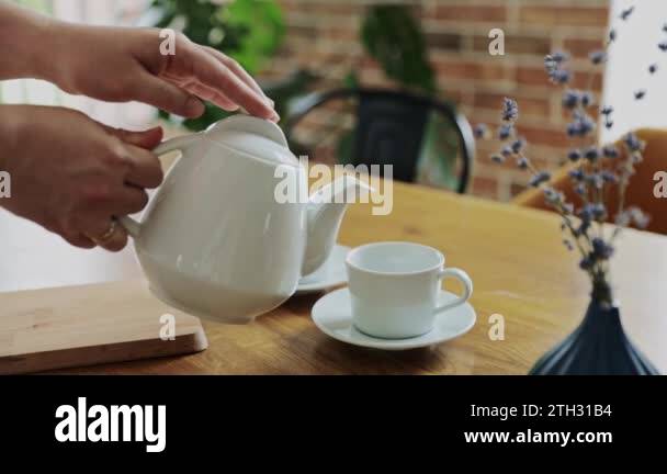 Hand is pouring hot drink from teapot into two tea cups, mugs indoors ...