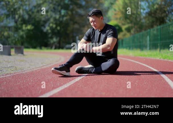 Asian runner massaging and rubbing injured leg while sitting on track ...