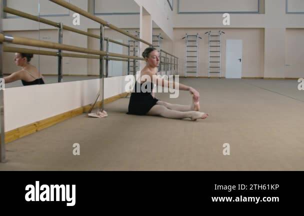 Ballet dancer in training suit near ballet barre stretches legs before practice at dance studio ...