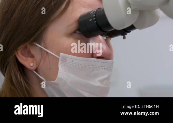 close-up face of female doctor looks into microscope in mask nose ...