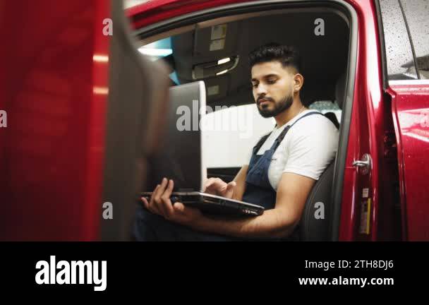 Bearded male mechanic sitting inside car using laptop, recording ...