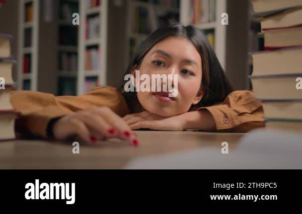 Tired pensive young asian woman lying on desk with books preparing for examination at university ...