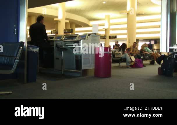 People sit and wait on benches in well lit airport terminal while airline employee looks at ...