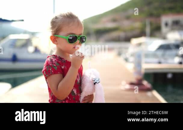 Little girl in sunglasses with a plush rabbit stands on the pier and ...