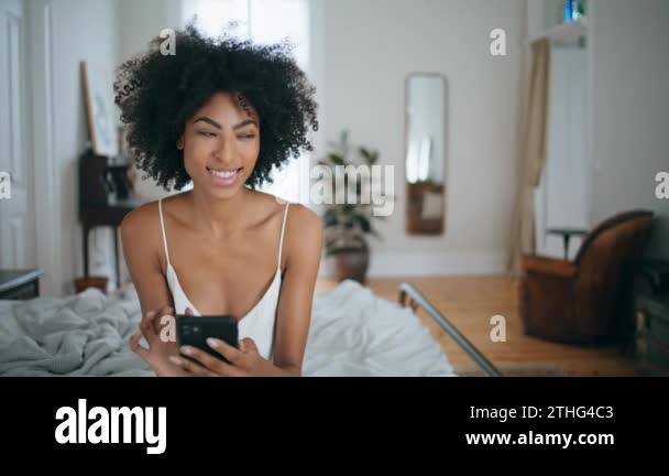 Smiling woman messaging smartphone at bedroom. Curly hair laughing lady texting sms using ...