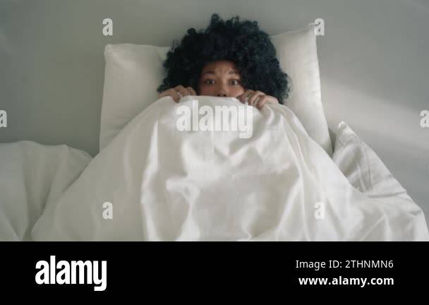 Scared african american woman peeking out from her blanket in bed with ...