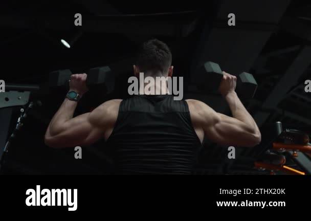 Athletic man back view lifting dumbbells over head in fitness gym ...