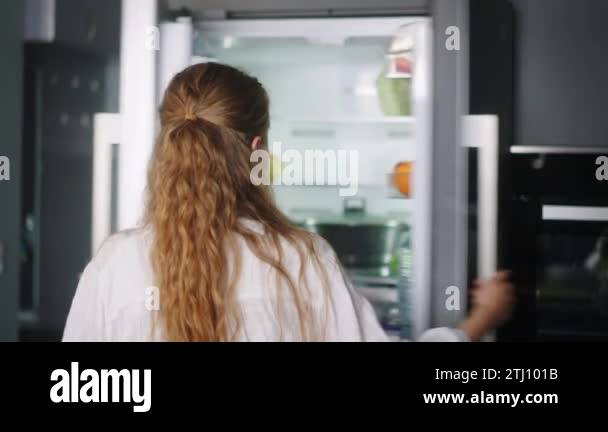 Young woman taking vegetables from fridge and has fun singing and ...