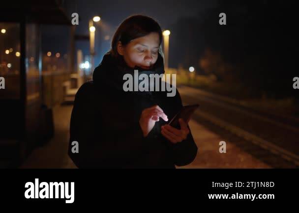A woman in black jacket on the platform of a city train at night using ...