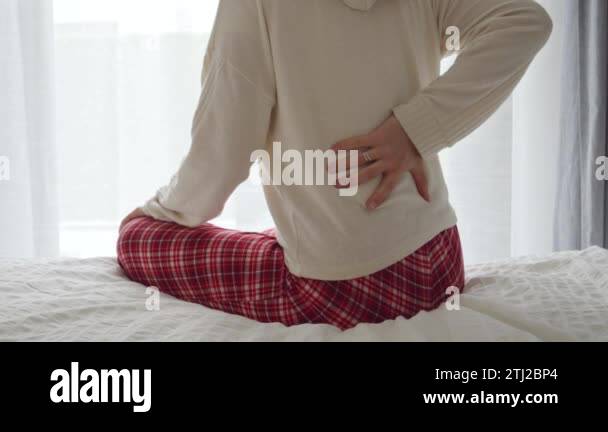 Young woman in pajamas sitting on the bed and holding her lower back ...