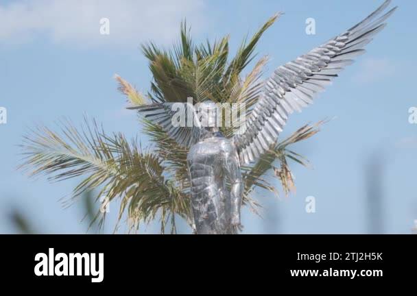 Winged man metal statue is standing in front of the palm on the street ...