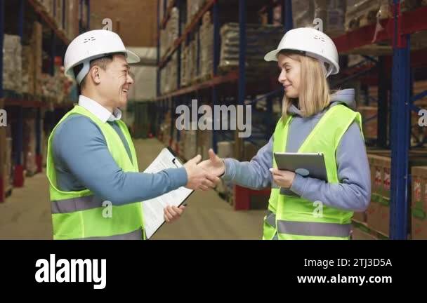 Multicultural male and female warehouse workers in white hardhats and reflective vests making ...